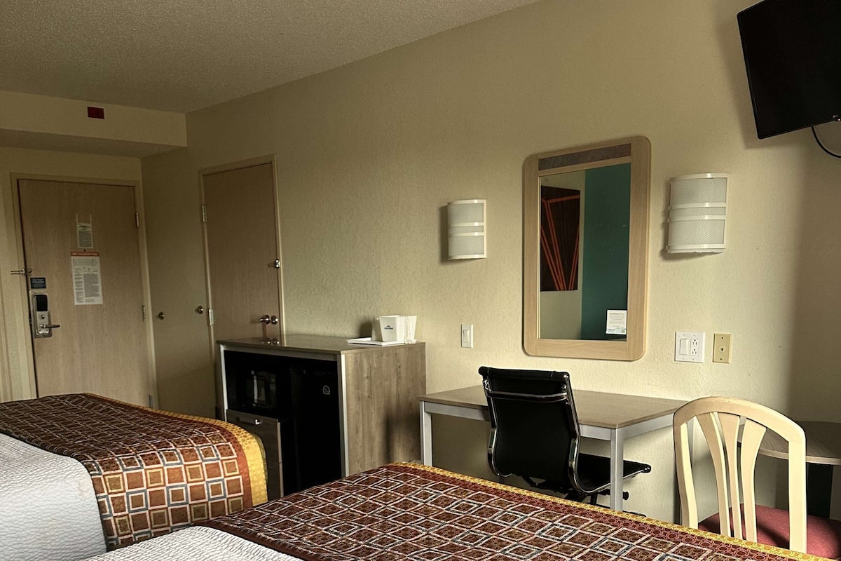 Quarto de hotel com duas camas, uma escrivaninha, uma cadeira, uma televisão e um espelho.