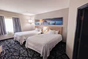 Quarto de hotel com duas camas, um quadro de um píer ao pôr do sol, uma mesa com abajur e uma janela com cortinas.