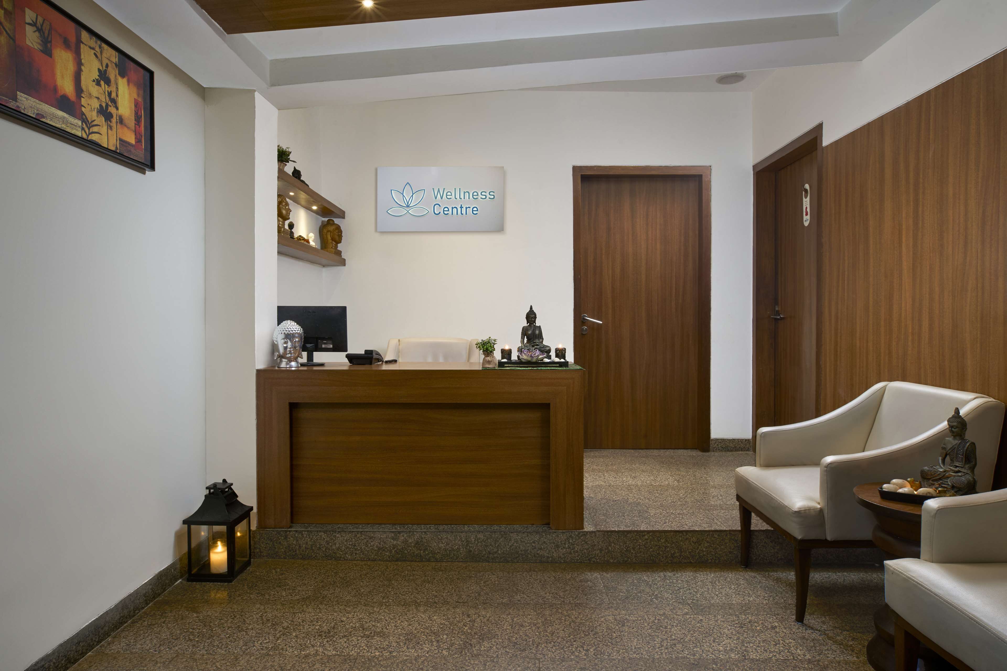 Uma área de recepção com uma mesa de madeira, uma placa com a inscrição "Wellness Centre" e uma estátua à direita.