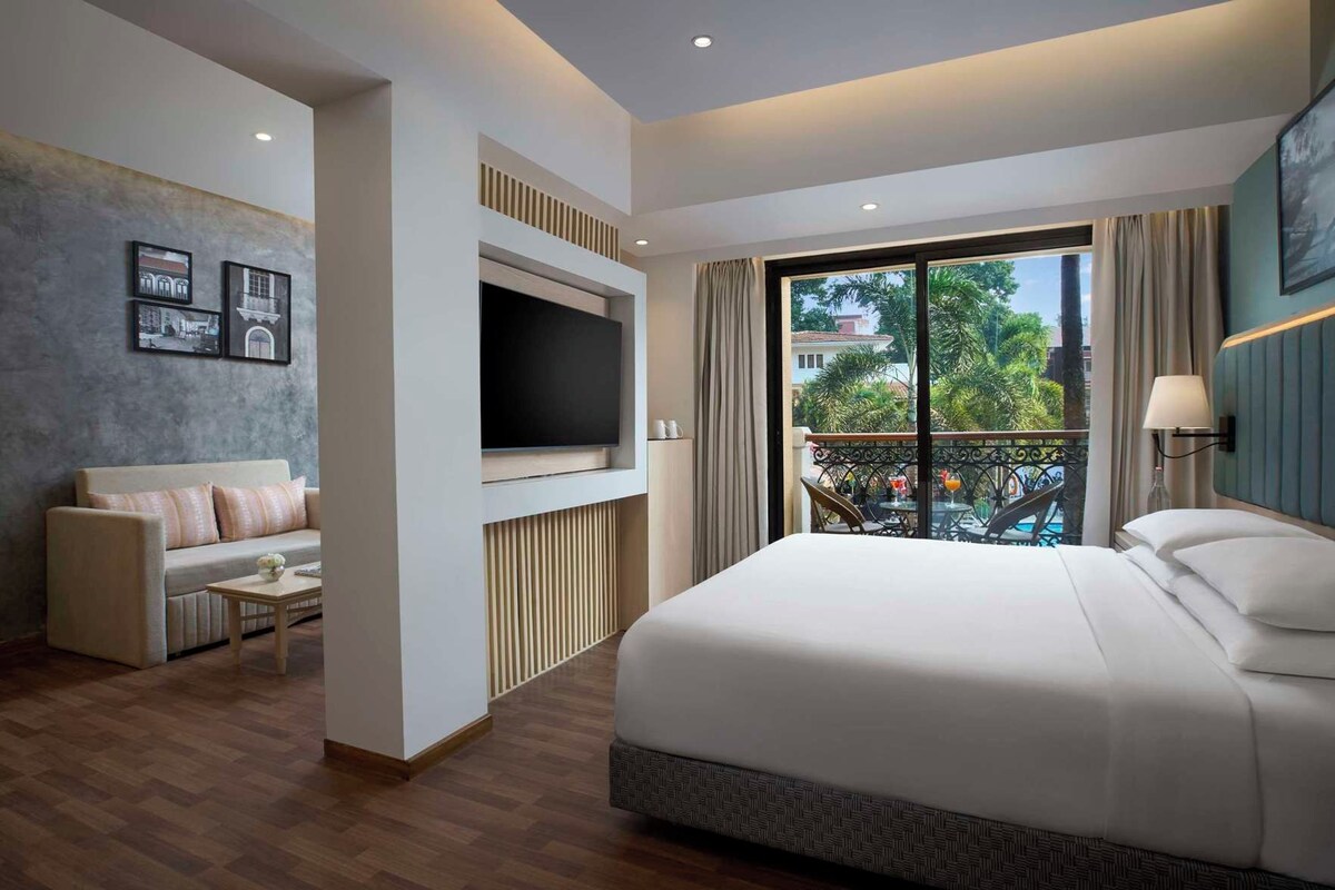 Quarto de hotel com cama grande, sofá, televisão na parede e varanda com vista para a cidade.