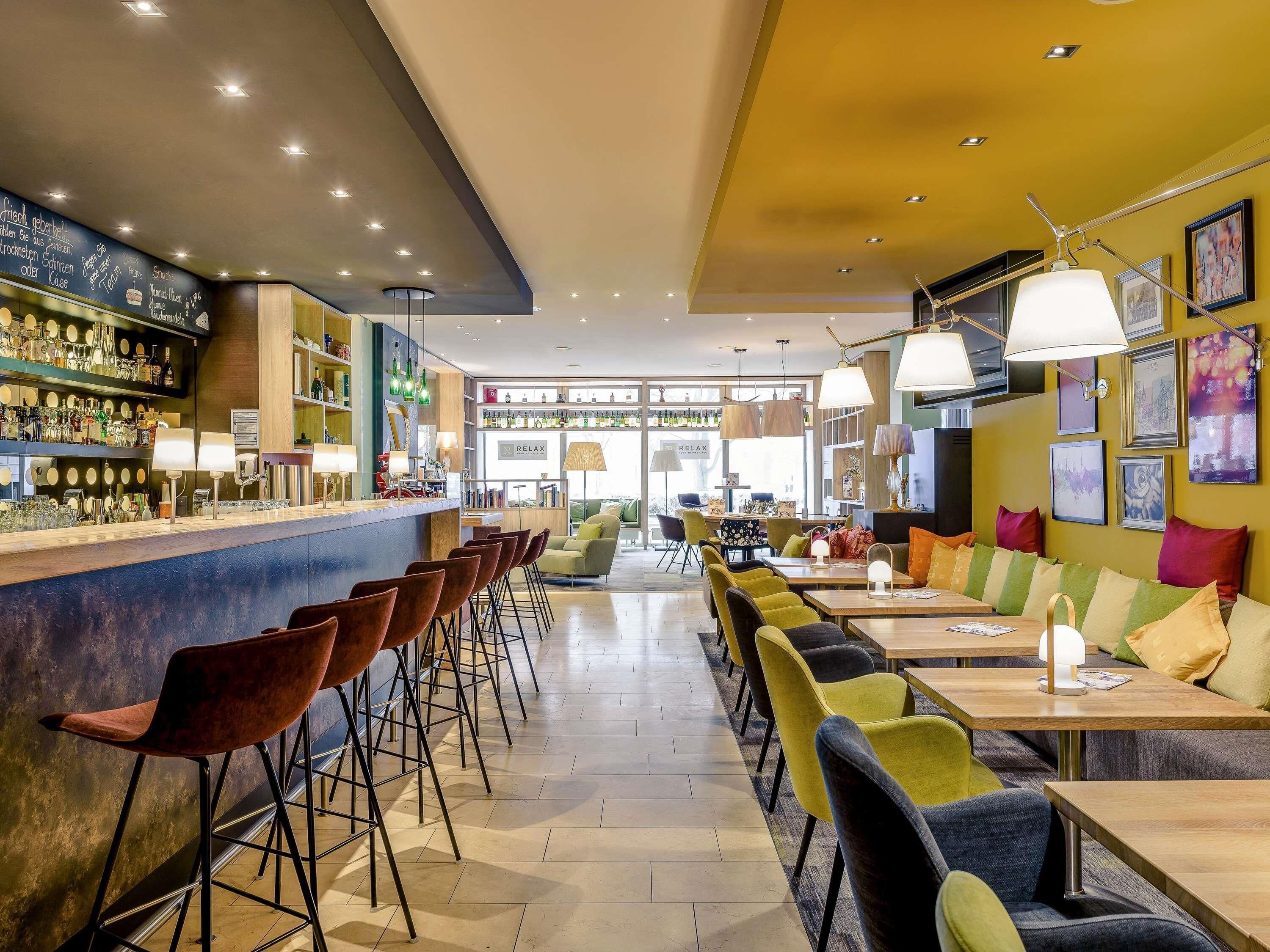 Uma área de bar moderna com bancos, balcão e prateleiras com garrafas. Há mesas e cadeiras na área de jantar, e um letreiro com a palavra 'RELAX'.