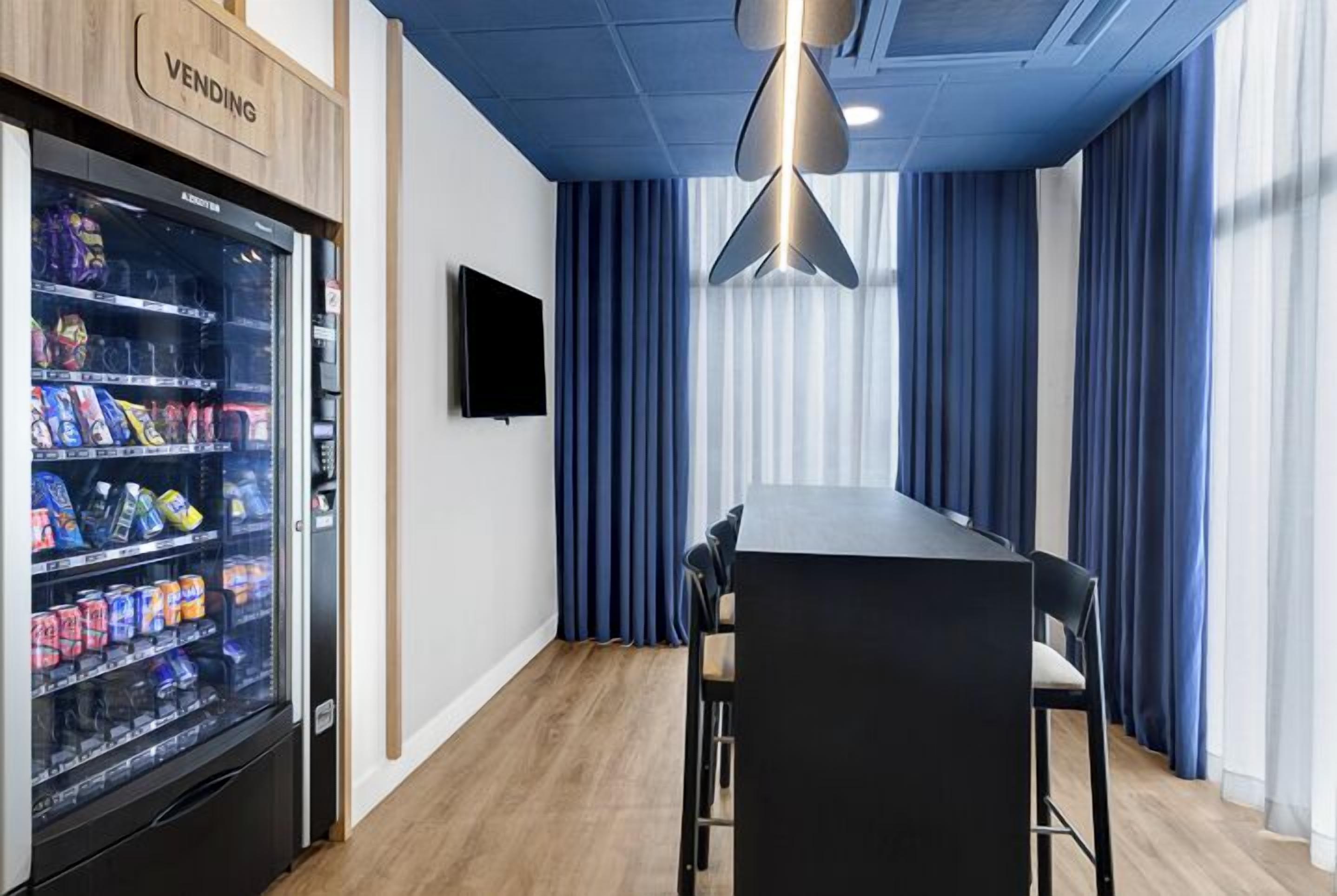 Quarto com máquina de venda automática, mesa, cadeiras, televisão e cortinas azuis.
