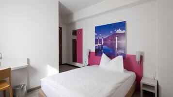 Quarto de hotel com cama, escrivaninha, cadeira, lixeira e um quadro na parede.