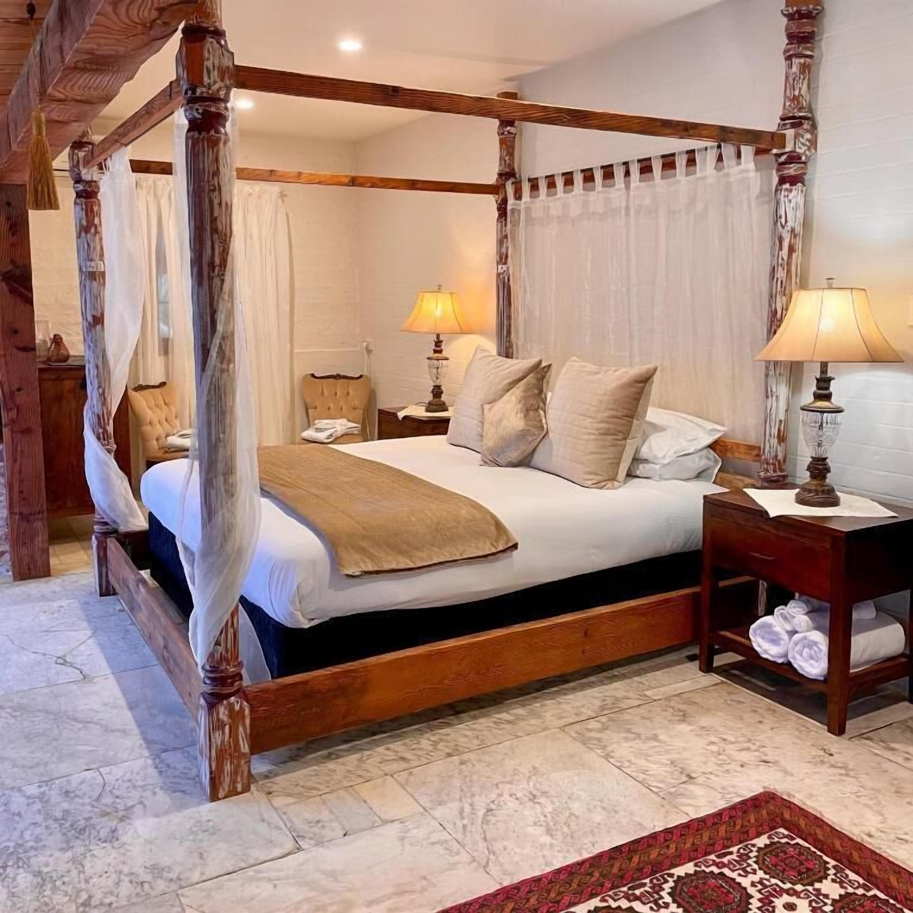 Uma cama de dossel com estrutura de madeira, uma mesinha de cabeceira com abajur e um tapete estampado no chão.