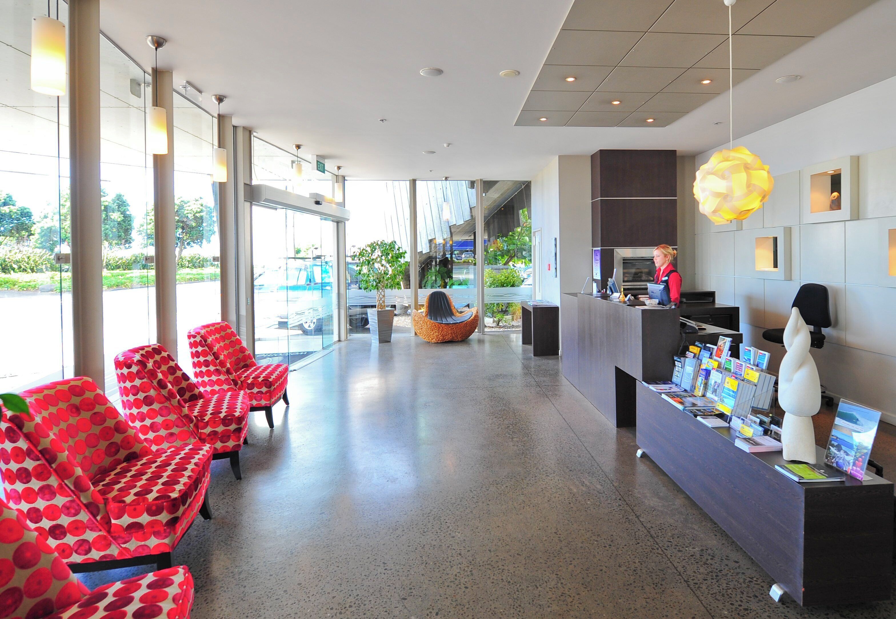 Um lobby moderno com cadeiras vermelhas de bolinhas, um balcão de recepção com folhetos e uma janela grande com vista para o exterior.