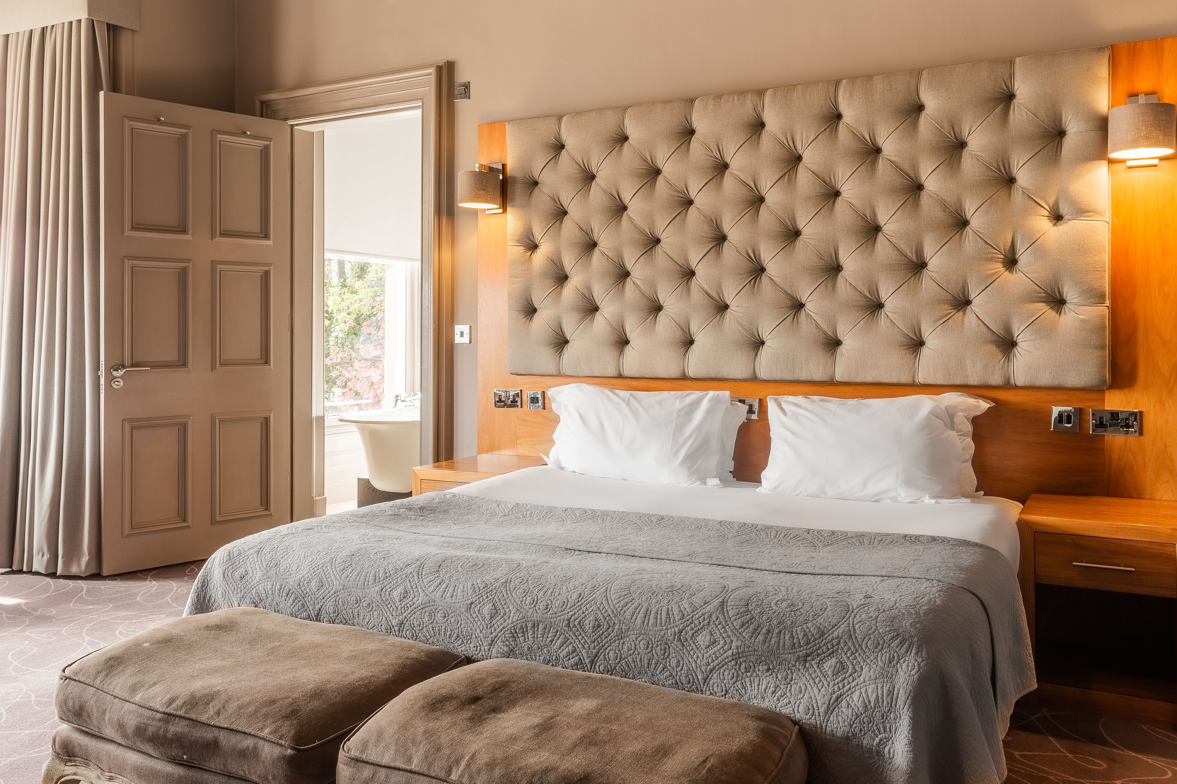 Quarto de hotel com uma cama grande, cabeceira estofada, mesa de cabeceira de madeira e uma janela com cortinas.
