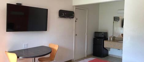 Quarto pequeno com mesa de jantar, duas cadeiras, TV de tela plana na parede, micro-ondas e geladeira no canto.