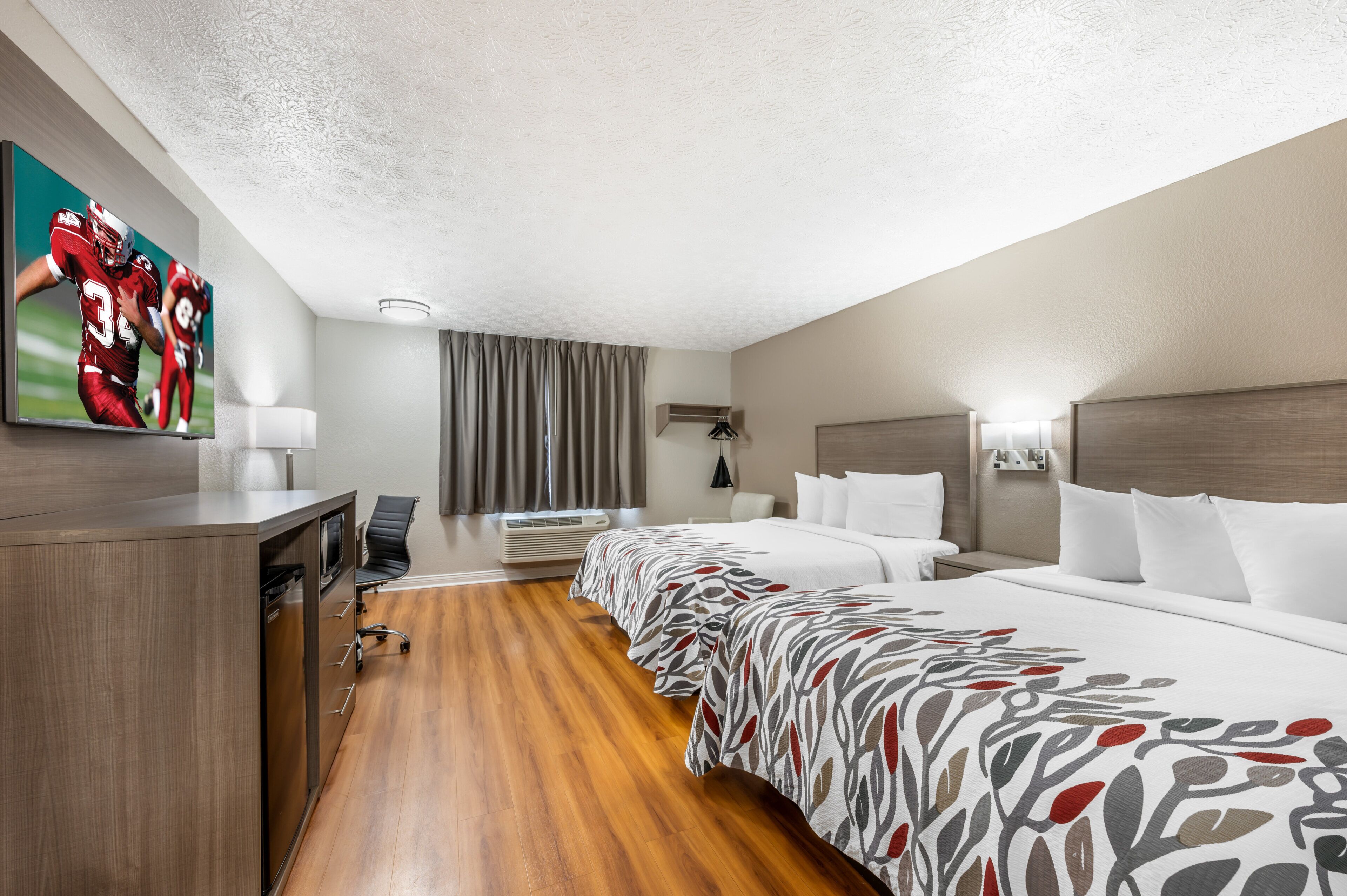 Quarto de hotel com piso de madeira, uma escrivaninha com cadeira, cama com roupa de cama branca, televisão na parede e janela com cortinas.