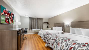 Quarto de hotel com piso de madeira, uma escrivaninha com cadeira, cama com roupa de cama branca, televisão na parede e janela com cortinas.