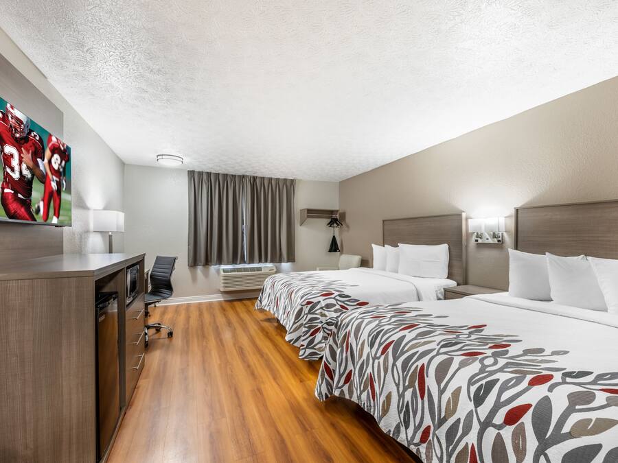 Quarto de hotel com piso de madeira, uma escrivaninha com cadeira, cama com roupa de cama branca, televisão na parede e janela com cortinas.