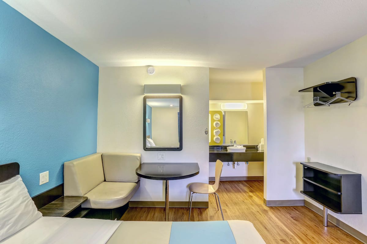 Quarto de hotel com cama, uma pequena escrivaninha, uma cadeira, um espelho, televisão e banheiro visível ao fundo.