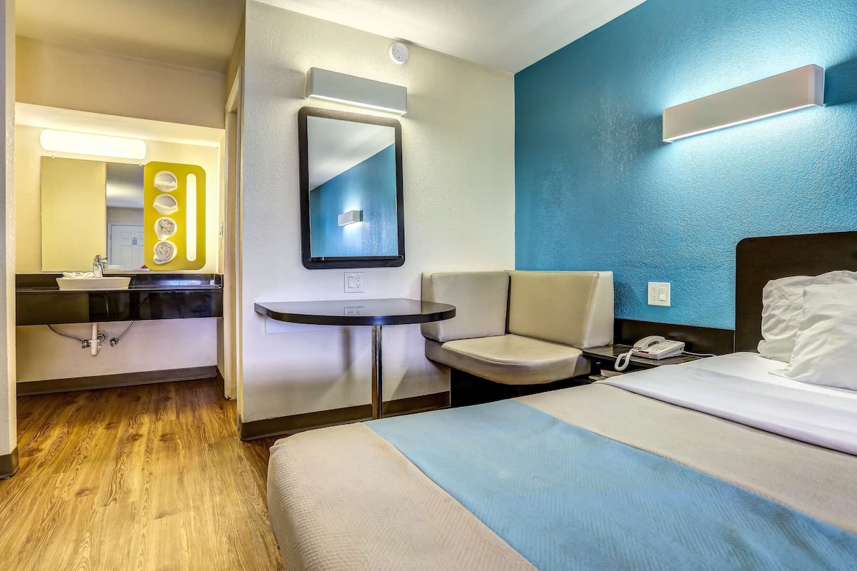 Quarto de hotel com cama, mesa de cabeceira, cadeira, mesinha e banheiro com pia e espelho.