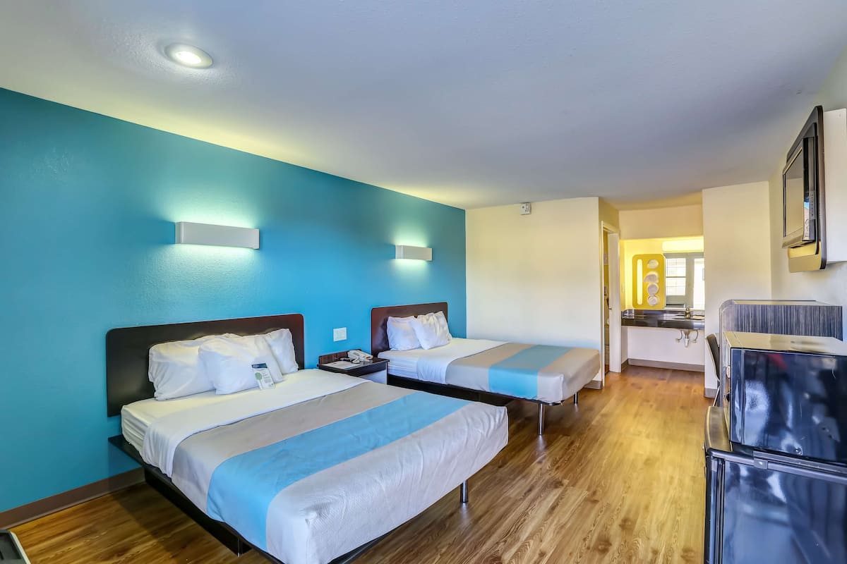 Quarto de hotel com duas camas, uma cozinha compacta e um banheiro visível ao fundo.