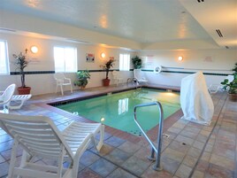 Piscina interna com piso ladrilhado, cadeiras de descanso brancas, plantas em vasos e placas de segurança nas paredes.