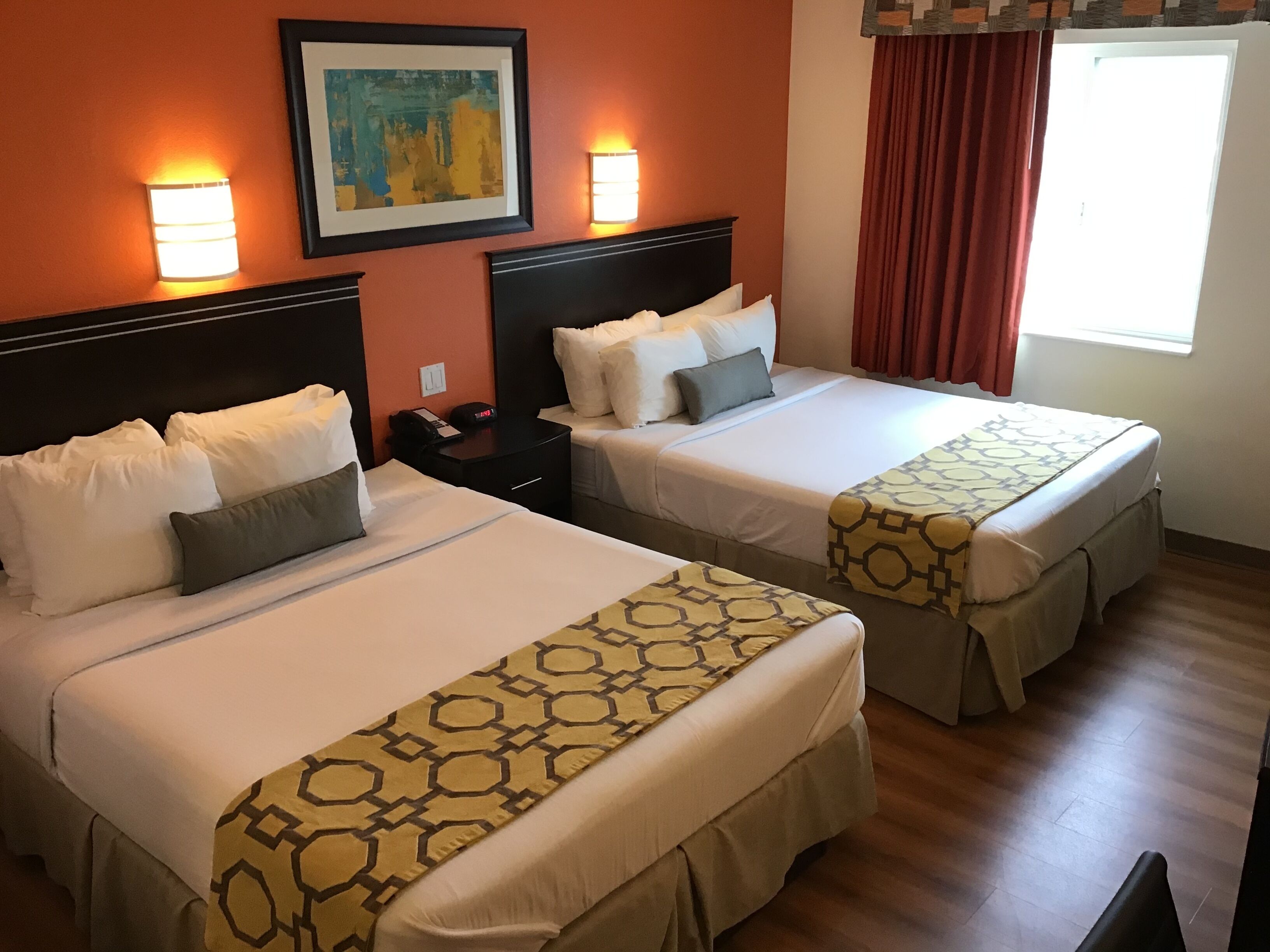 Quarto de hotel com duas camas, cada uma com colcha estampada, uma mesa de cabeceira, um quadro pendurado na parede e uma janela com cortinas vermelhas.