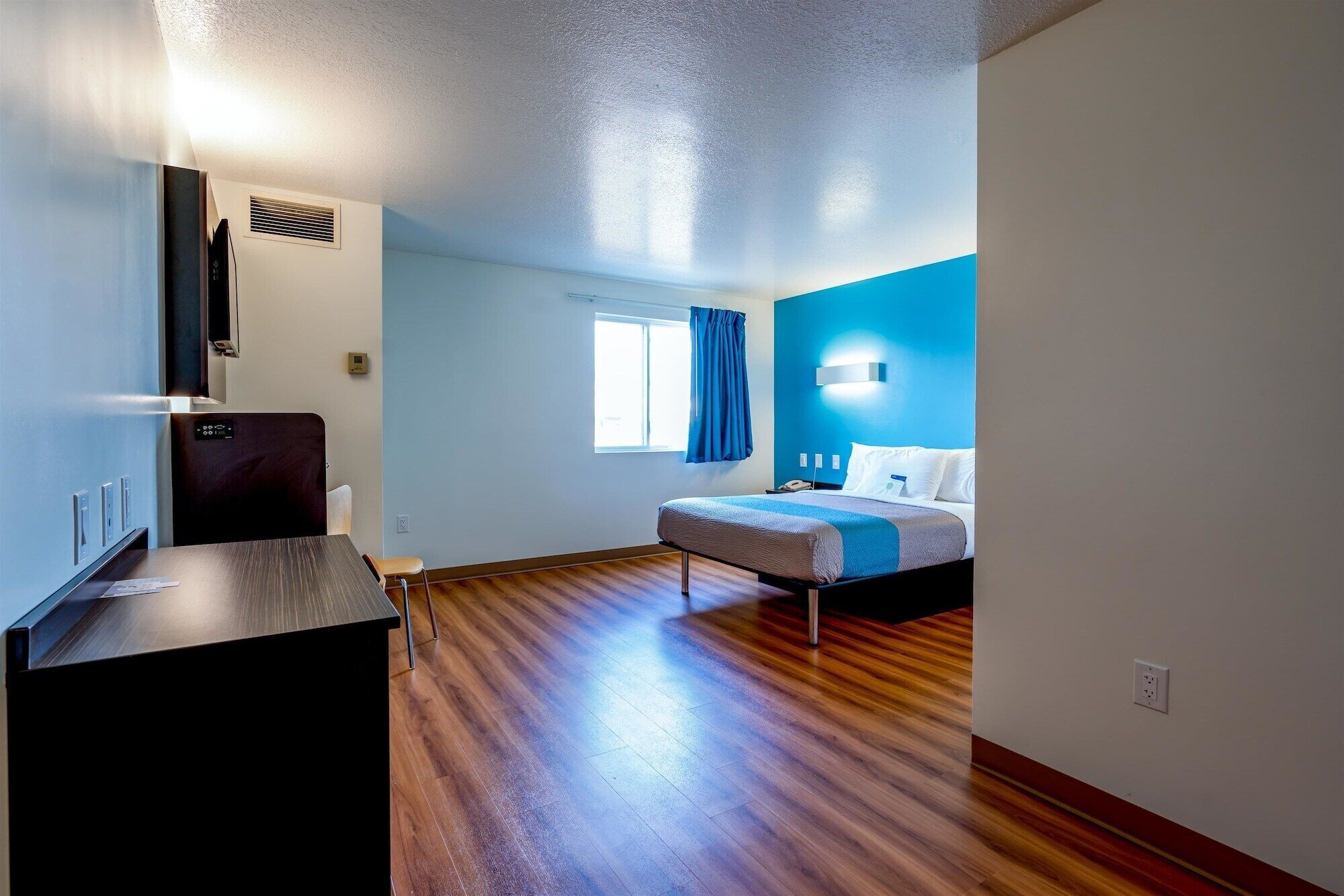 Quarto de hotel com piso de madeira, cama com parede azul decorativa, escrivaninha com cadeira e janela com cortinas.