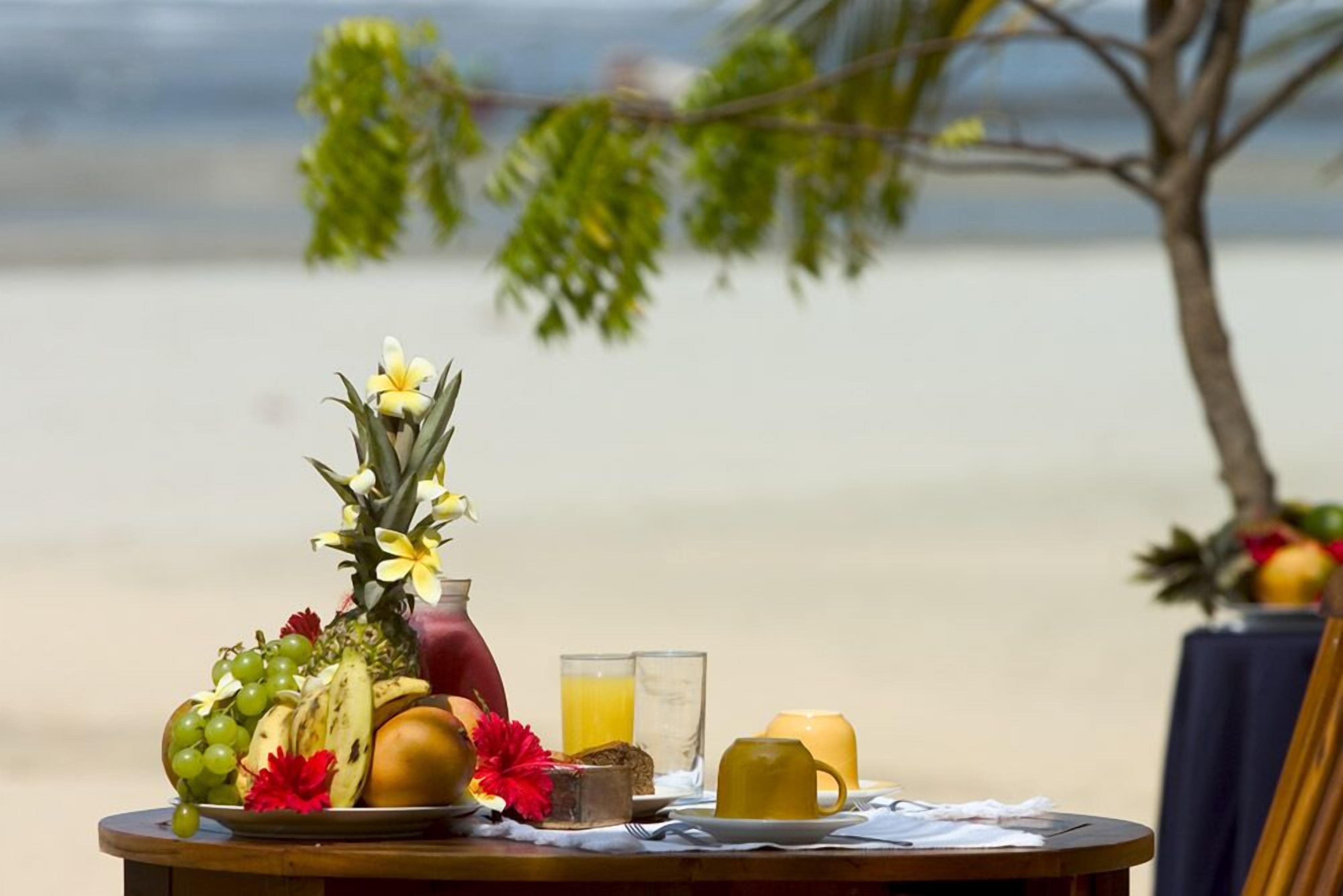 Uma mesa com uma travessa de frutas, um vaso com flores, um copo de suco de laranja e uma caneca em um ambiente praiano.