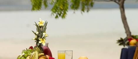 Uma mesa com uma travessa de frutas, um vaso com flores, um copo de suco de laranja e uma caneca em um ambiente praiano.