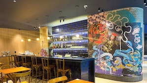 Uma área de bar com um mural, mesas e cadeiras de madeira e piso ladrilhado.