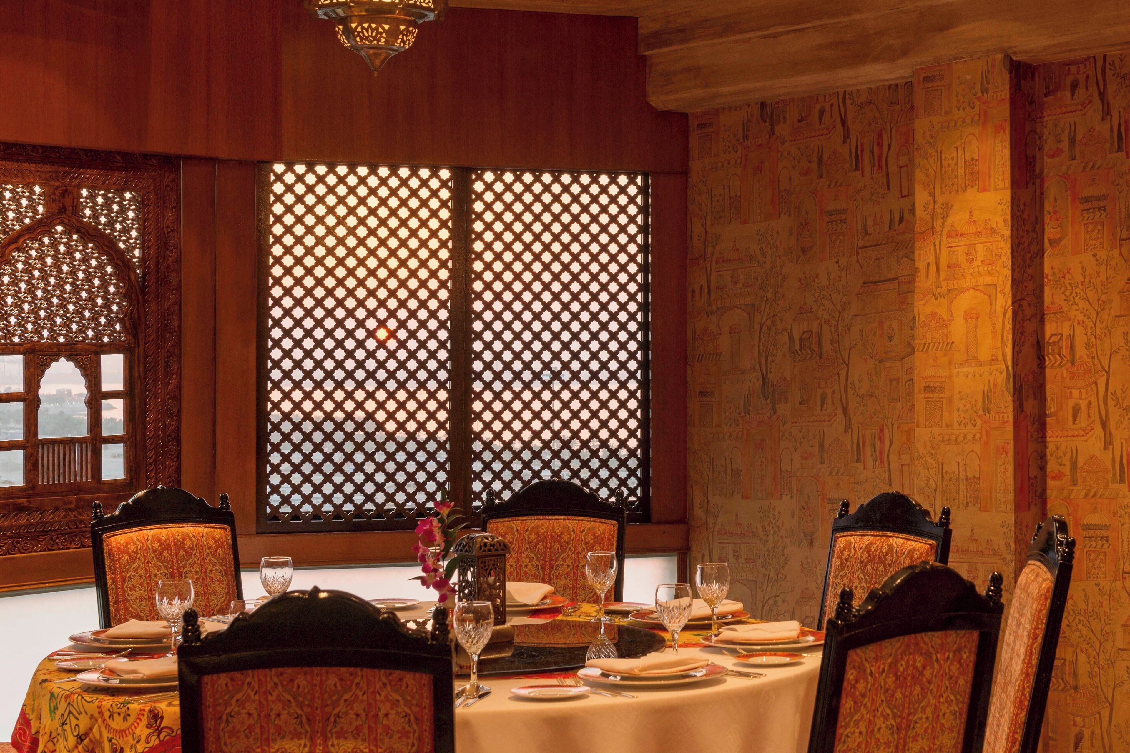 Uma sala de jantar com uma mesa posta para quatro pessoas, cadeiras elaboradas e uma janela decorativa com padrão em treliça.