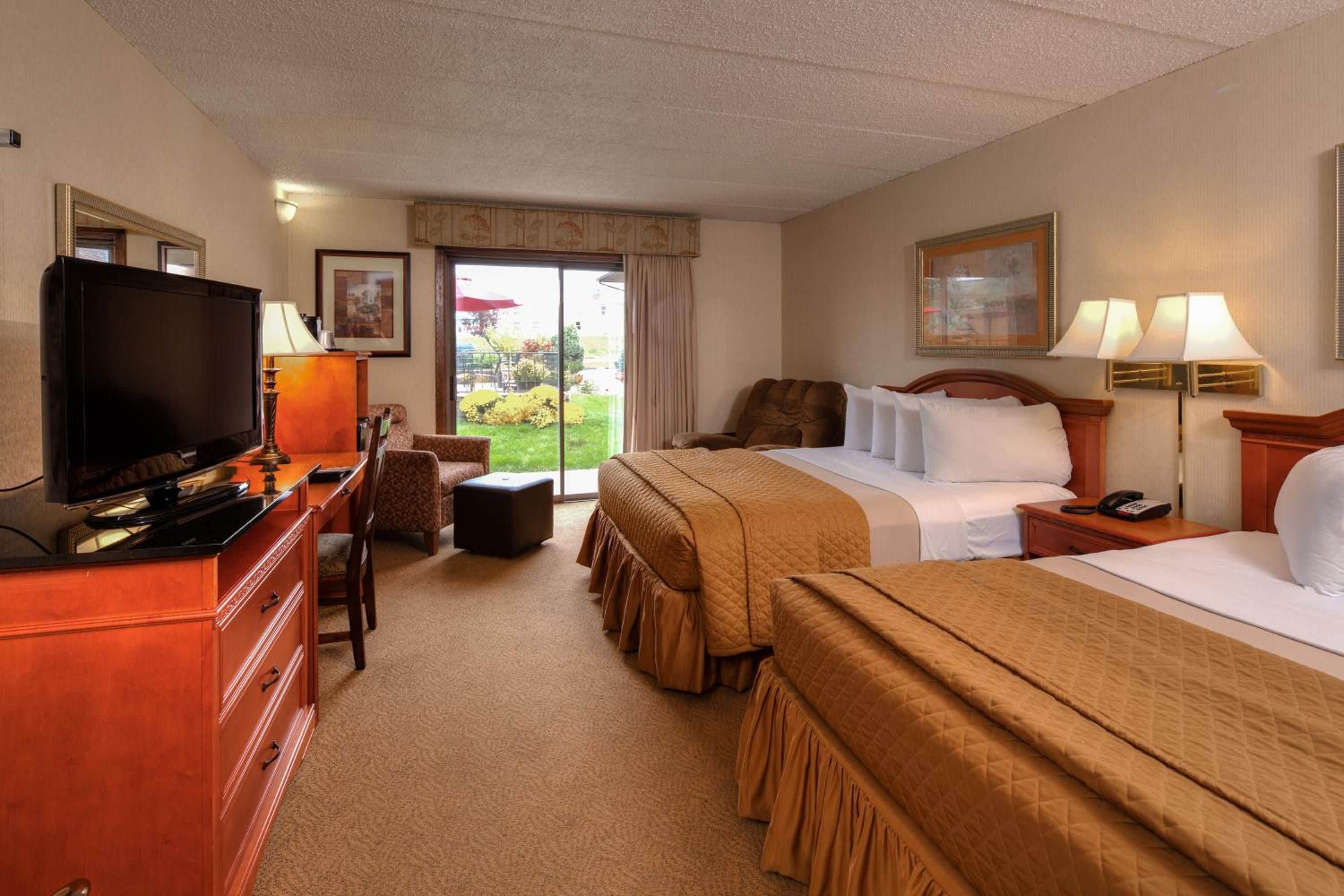 Quarto de hotel com televisão, escrivaninha, cadeira, cama com duas almofadas, mesa de cabeceira com abajur e janela com vista para o exterior.