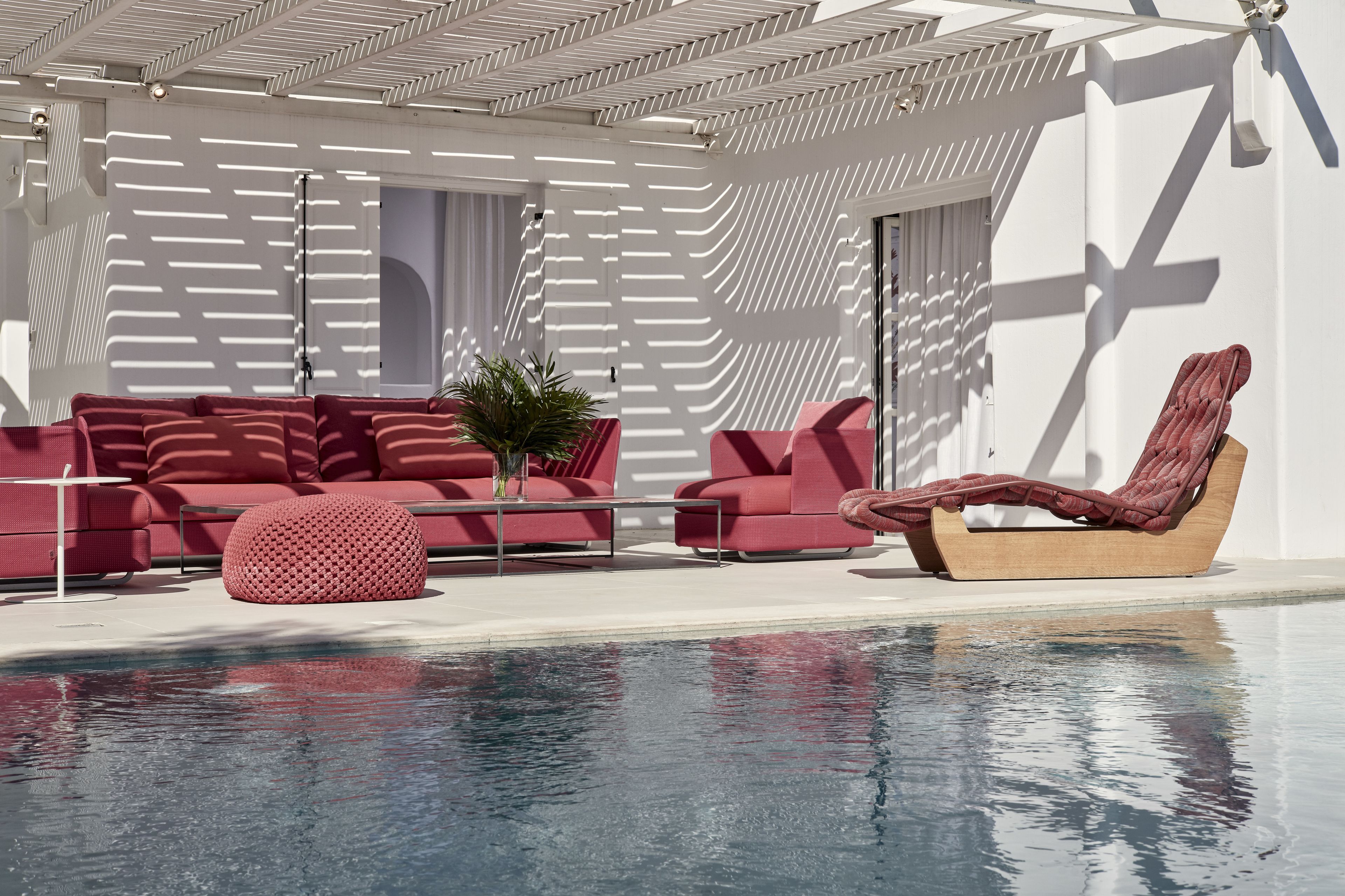 Uma área de piscina ao ar livre moderna com móveis vermelhos, incluindo uma espreguiçadeira e um banquinho, e um vaso com planta.