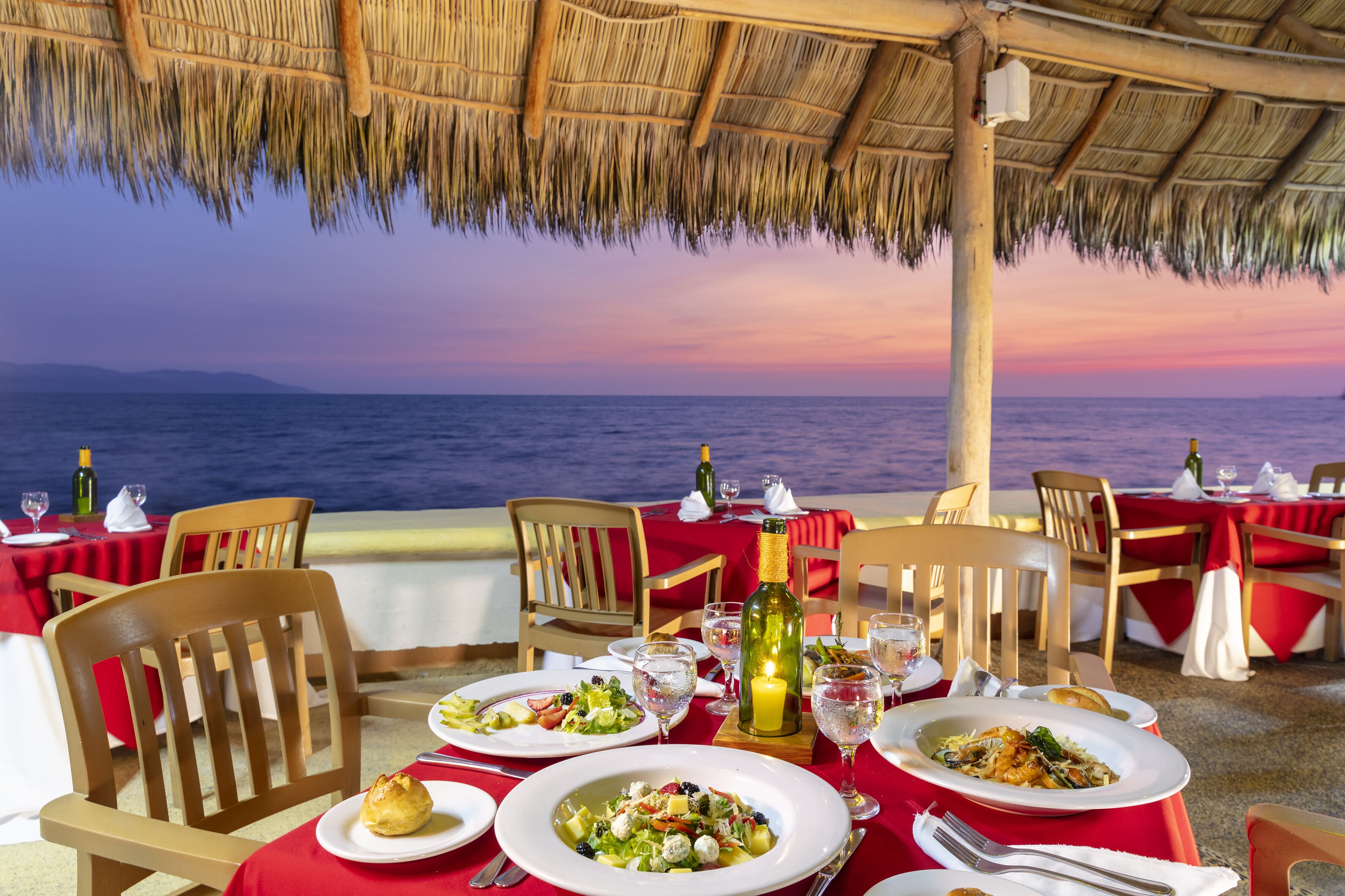 Mesa posta para o jantar com vista para o mar, cadeiras de madeira e um telhado de palha.