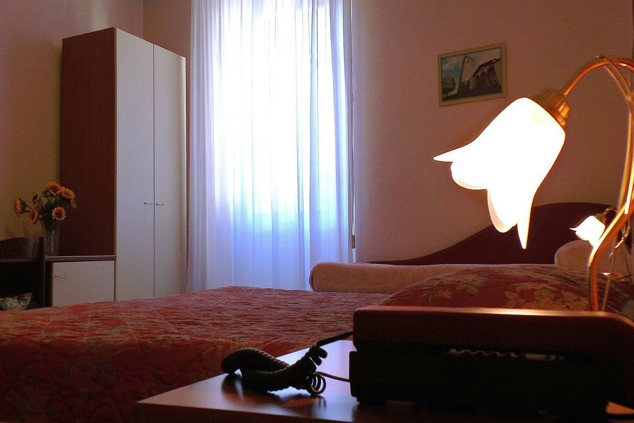 Quarto de hotel com cama, mesa de cabeceira, abajur, guarda-roupa e janela com cortinas.