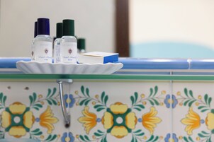 Casa de banho com painel de azulejos decorativo, uma pia branca e três frascos de amenidades de hotel sobre um tabuleiro.
