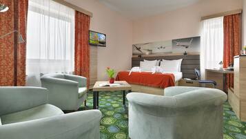 Quarto de hotel com cama, duas poltronas, uma escrivaninha, televisão e janela com cortinas.