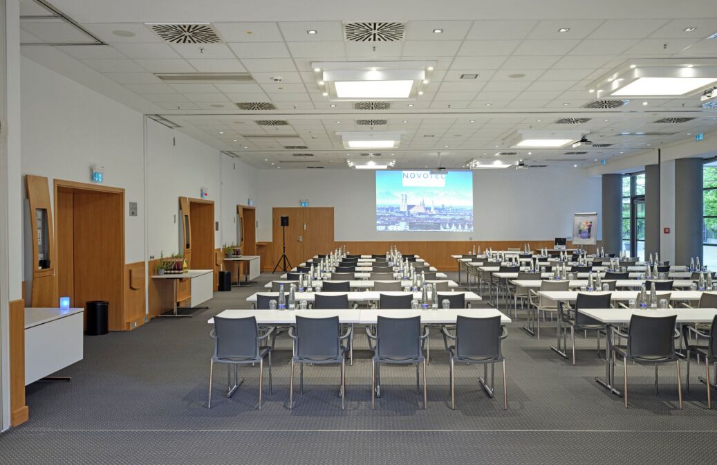Uma sala de conferências com várias mesas e cadeiras, uma tela de projeção e uma imagem de paisagem urbana na parede.