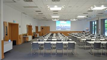 Uma sala de conferências com várias mesas e cadeiras, uma tela de projeção e uma imagem de paisagem urbana na parede.