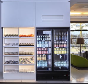 Um refrigerador de bebidas repleto de diversas opções, incluindo água, refrigerante e cerveja, em um ambiente moderno com vista para o exterior.