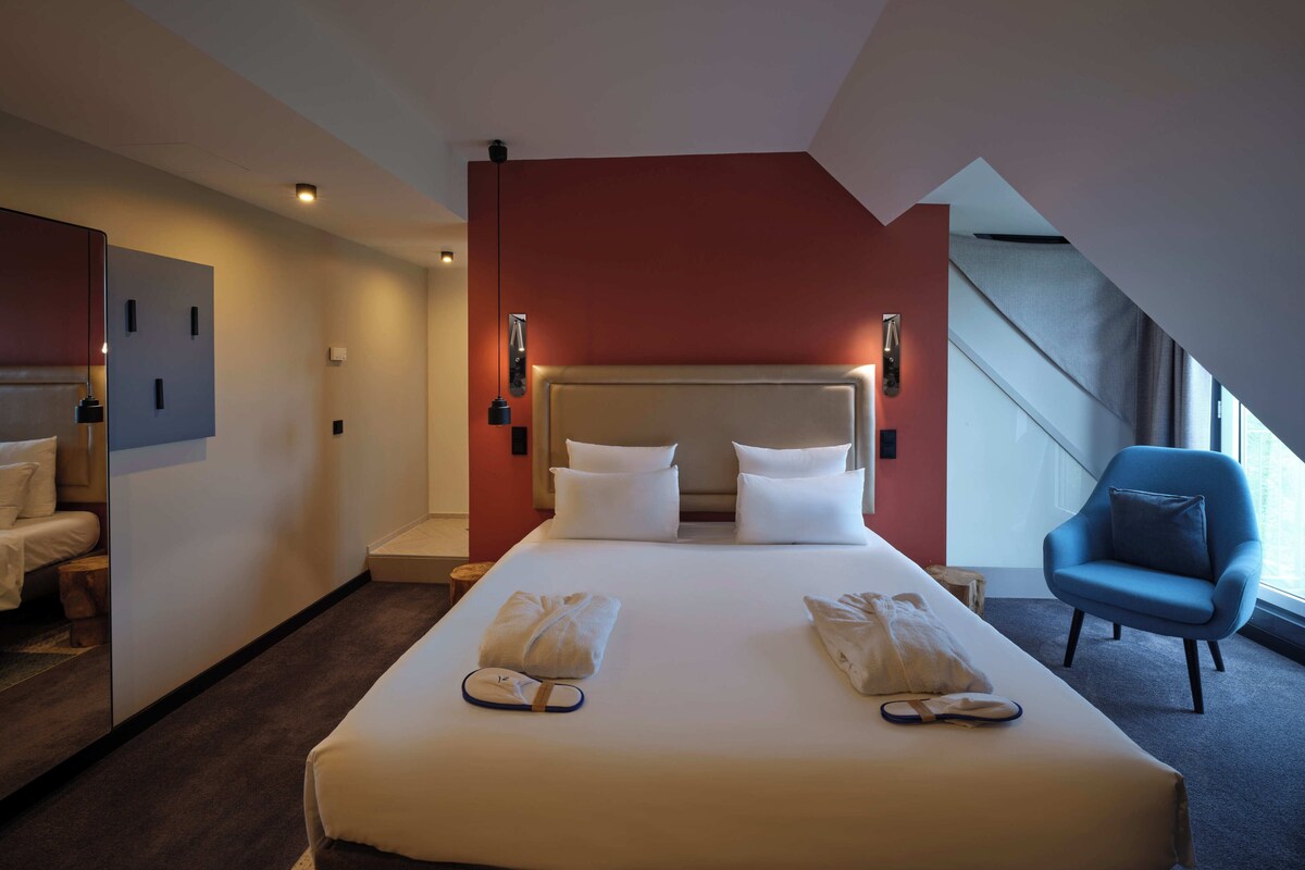Quarto de hotel com cama grande, duas mesas de cabeceira, uma poltrona azul e vista para o exterior.