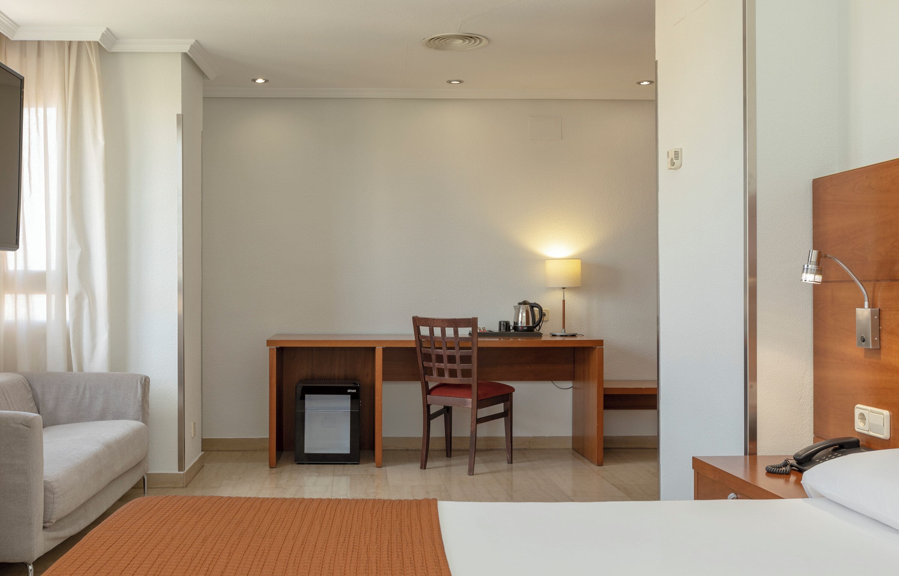 Quarto de hotel com cama, escrivaninha com cadeira, mini-geladeira, luminária e mesa de cabeceira com telefone.