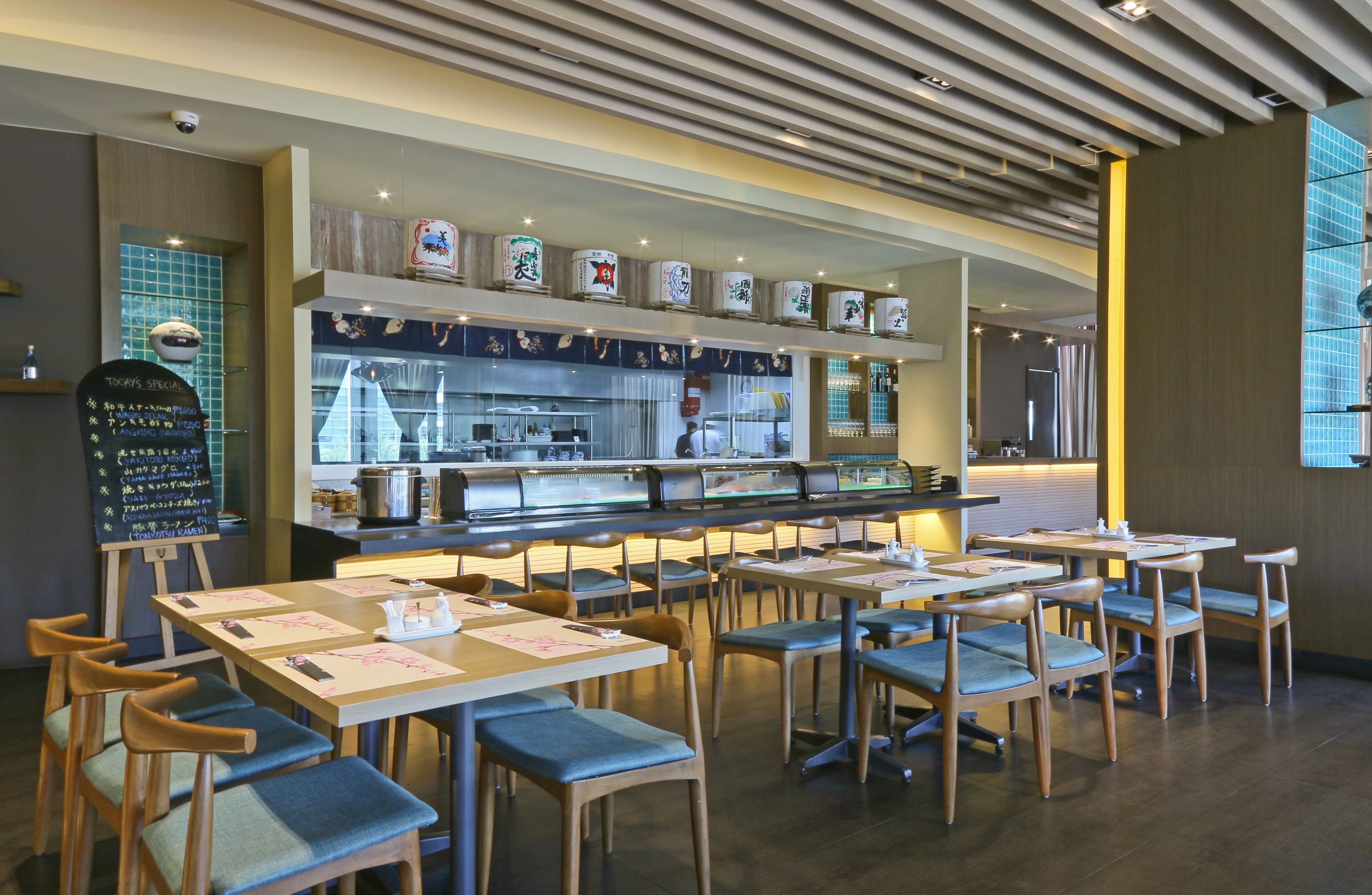 Um restaurante com várias mesas e cadeiras, um balcão com vitrine e um menu em lousa.