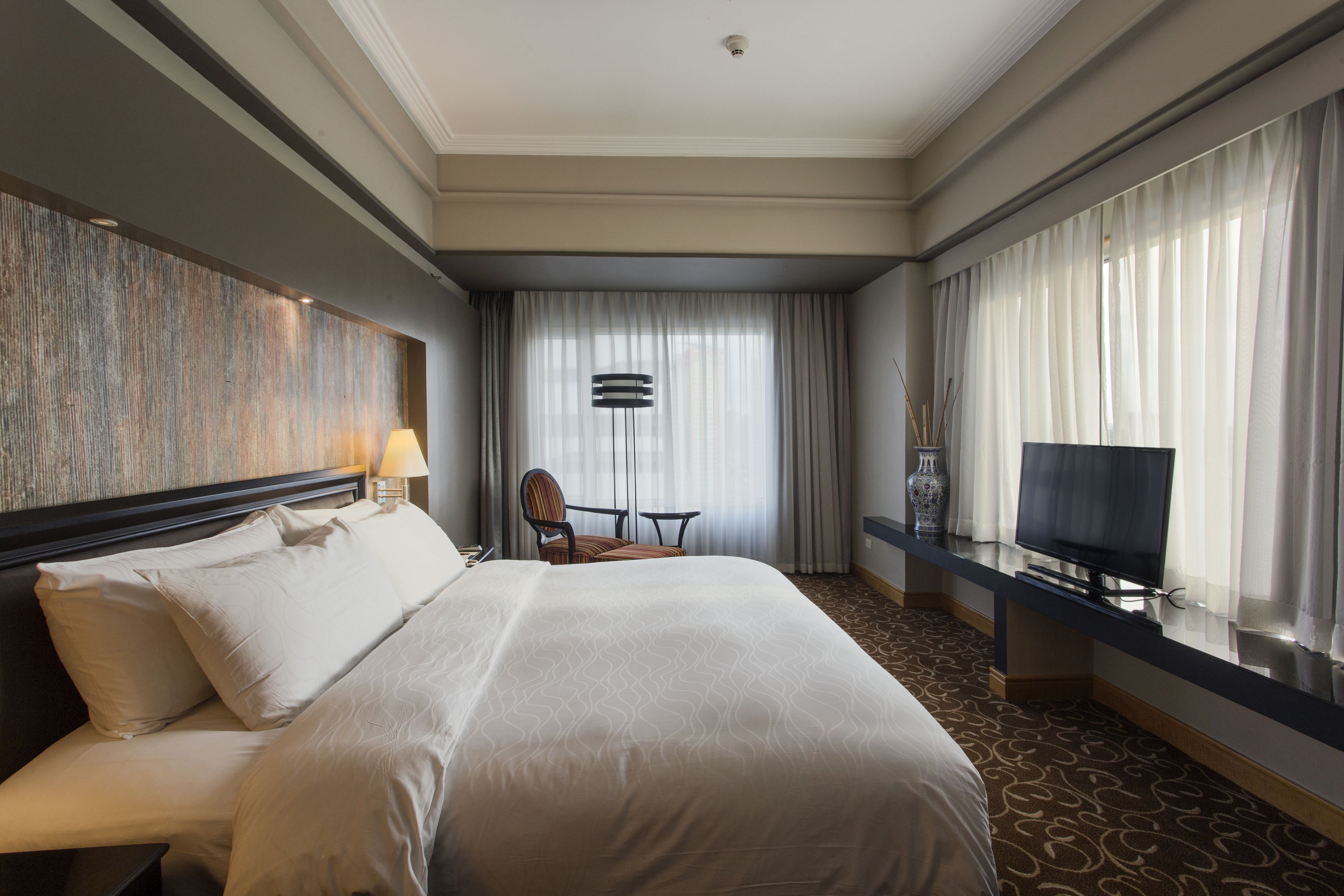Quarto de hotel com uma cama grande, televisão em um suporte, uma cadeira e uma escrivaninha com abajur.