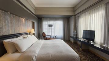 Quarto de hotel com uma cama grande, televisão em um suporte, uma cadeira e uma escrivaninha com abajur.