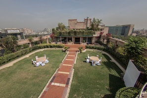 Um jardim no terraço com um caminho central, duas mesas redondas com cadeiras e um edifício com telhado verde ao fundo.
