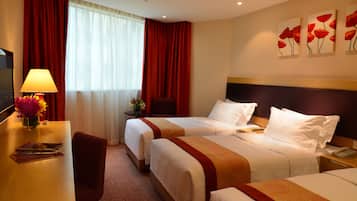 Quarto de hotel com duas camas, uma escrivaninha com luminária, uma cadeira e uma janela com cortinas.