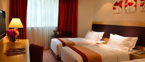 Quarto de hotel com duas camas, uma escrivaninha com luminária, uma cadeira e uma janela com cortinas.