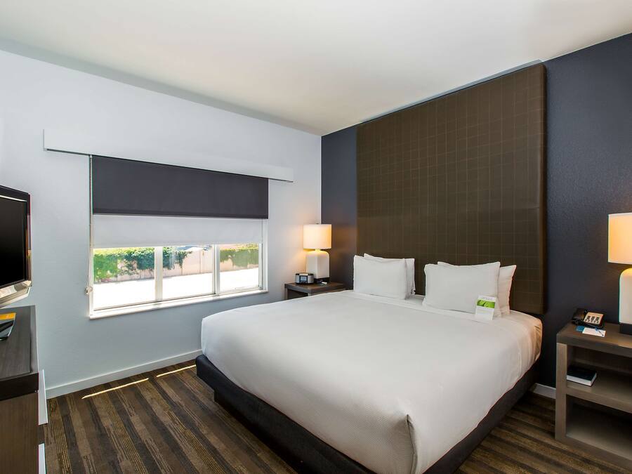 Quarto de hotel com uma cama grande, duas mesas de cabeceira com luminárias, uma TV de tela plana e uma janela com vista para áreas verdes.