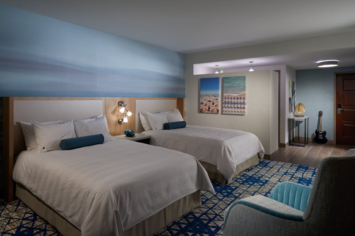 Quarto de hotel com duas camas, um tapete estampado, uma cadeira e obras de arte nas paredes.