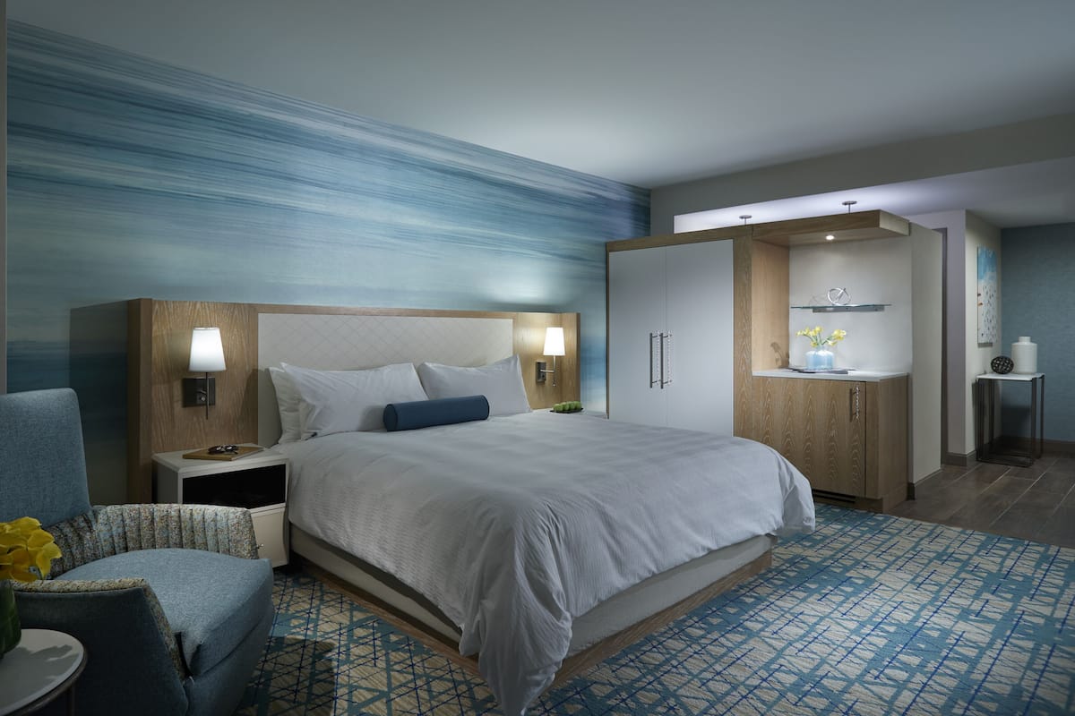 Quarto de hotel moderno com uma cama grande, área de estar com uma poltrona, uma pequena mesa com um vaso de flores e um guarda-roupa com espelho.