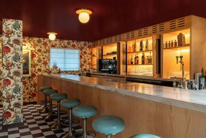 Uma área de bar com piso xadrez, balcão de madeira, banquetas e diversas garrafas e copos.