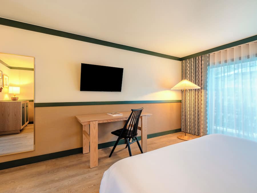 Quarto de hotel com cama, escrivaninha, cadeira, televisão e janela com cortinas.