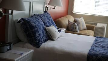 Cama bem arrumada com almofadas estampadas, luminária de cabeceira e mesa pequena com telefone e papéis.