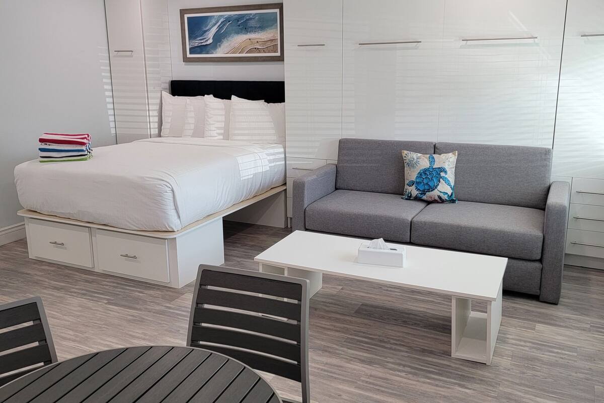 Quarto moderno com cama, sofá cinzento, secretária branca e mesa de jantar com cadeiras.