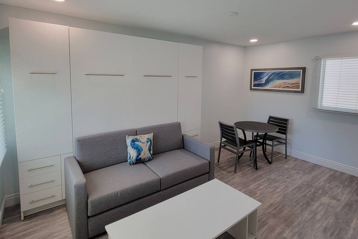 Uma sala de estar moderna com um sofá cinzento, uma mesa de centro branca, uma mesa de jantar com cadeiras e um guarda-roupa grande.