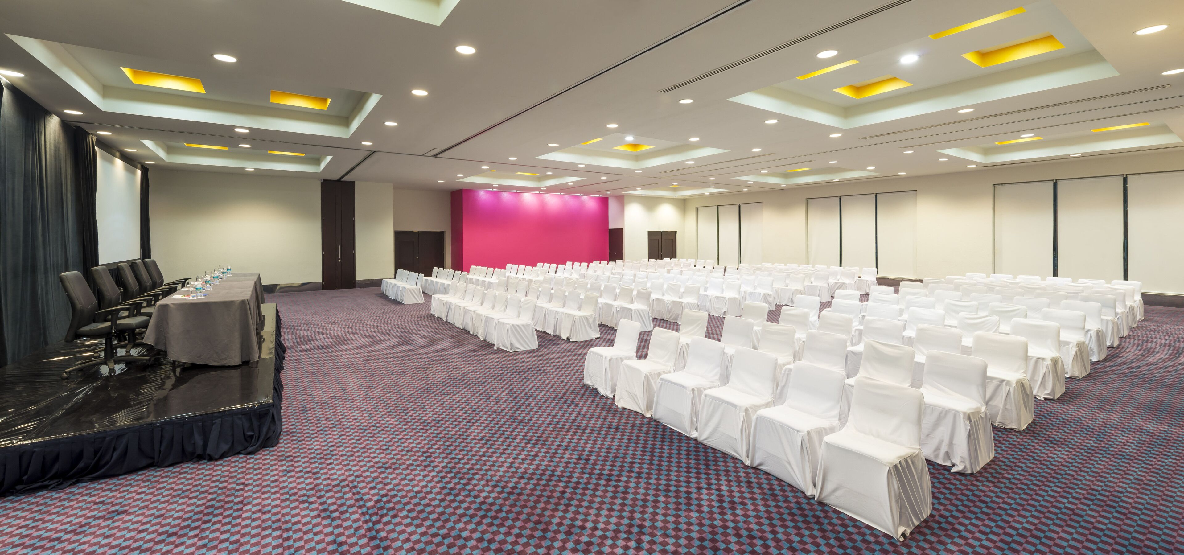 Uma sala de conferências com palco, fileiras de cadeiras brancas e um tapete estampado.